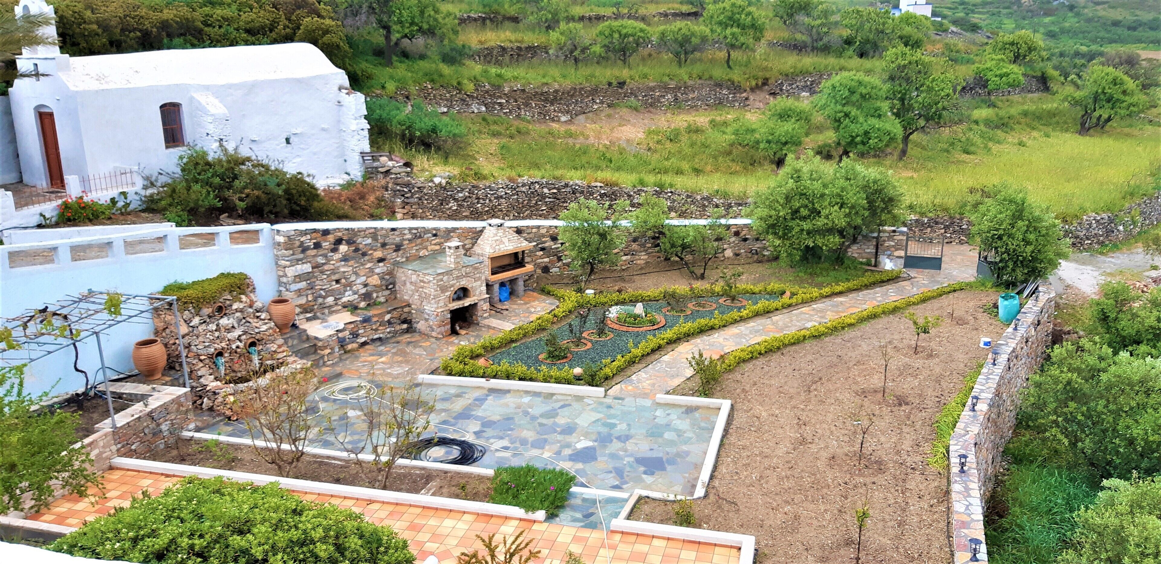 Um edifício branco com um pátio de pedra, piscina em mosaico e jardim com árvores e uma fonte.