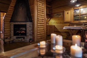 Interior de uma cabana de madeira com lareira de pedra, paredes de madeira e uma mesa com velas.