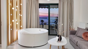 Uma sala de estar moderna com uma grande banheira redonda, um sofá cinza, uma pequena mesa branca e vista para o oceano através de uma porta de vidro.