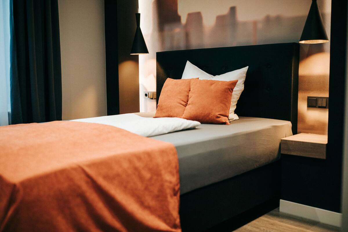 Quarto de hotel moderno com uma cama bem arrumada, mesa de cabeceira com abajur e vista para a cidade.