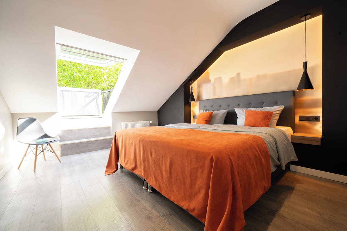 Quarto moderno com teto inclinado, uma janela grande, uma cama com colcha laranja e uma cadeira.