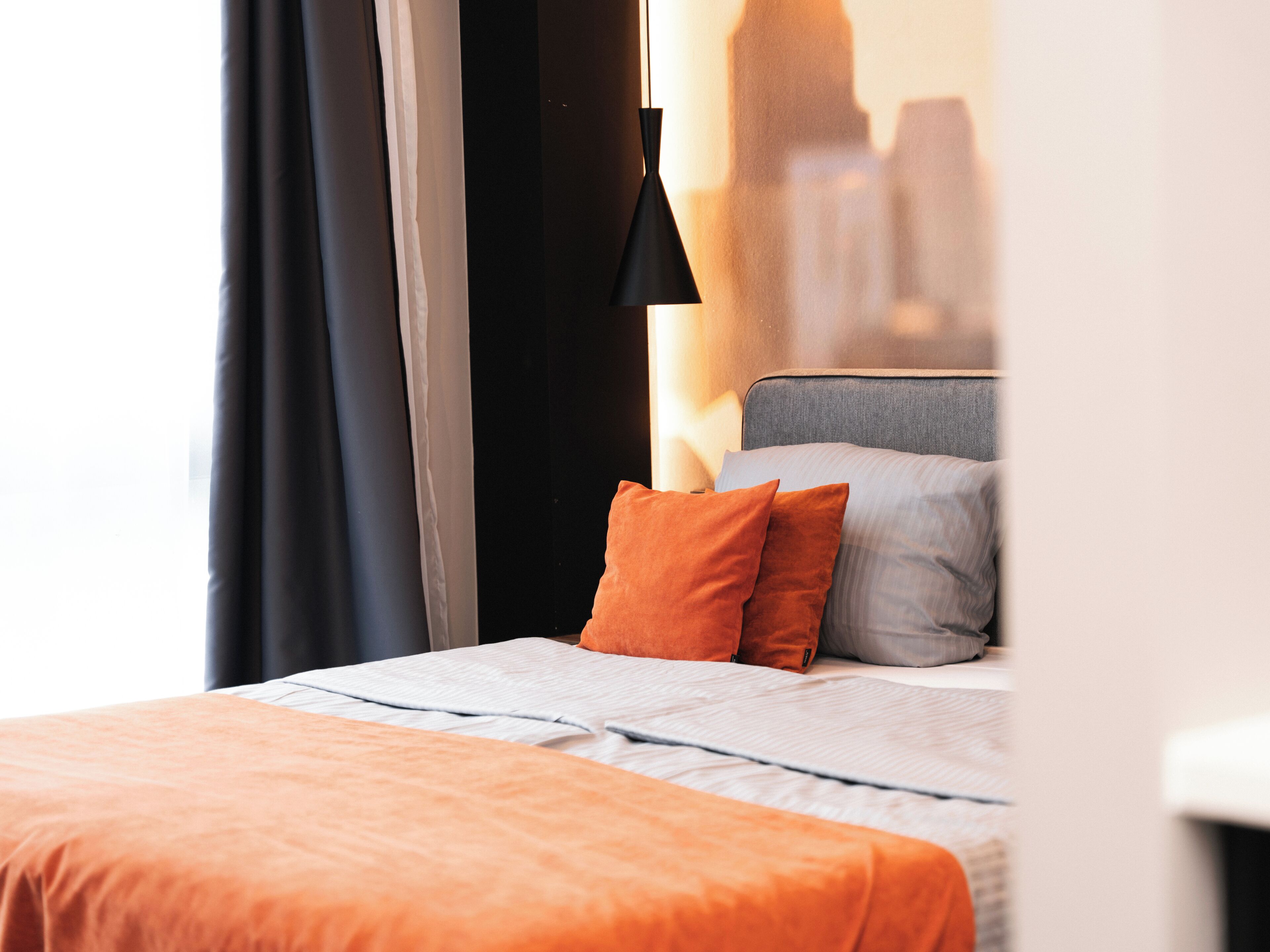 Cama arrumada com manta e almofadas laranja, abajur preto e vista para a cidade através da janela.
