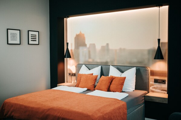 Quarto moderno com vista para a cidade, uma cama com roupa de cama laranja e duas luminárias pendentes pretas.