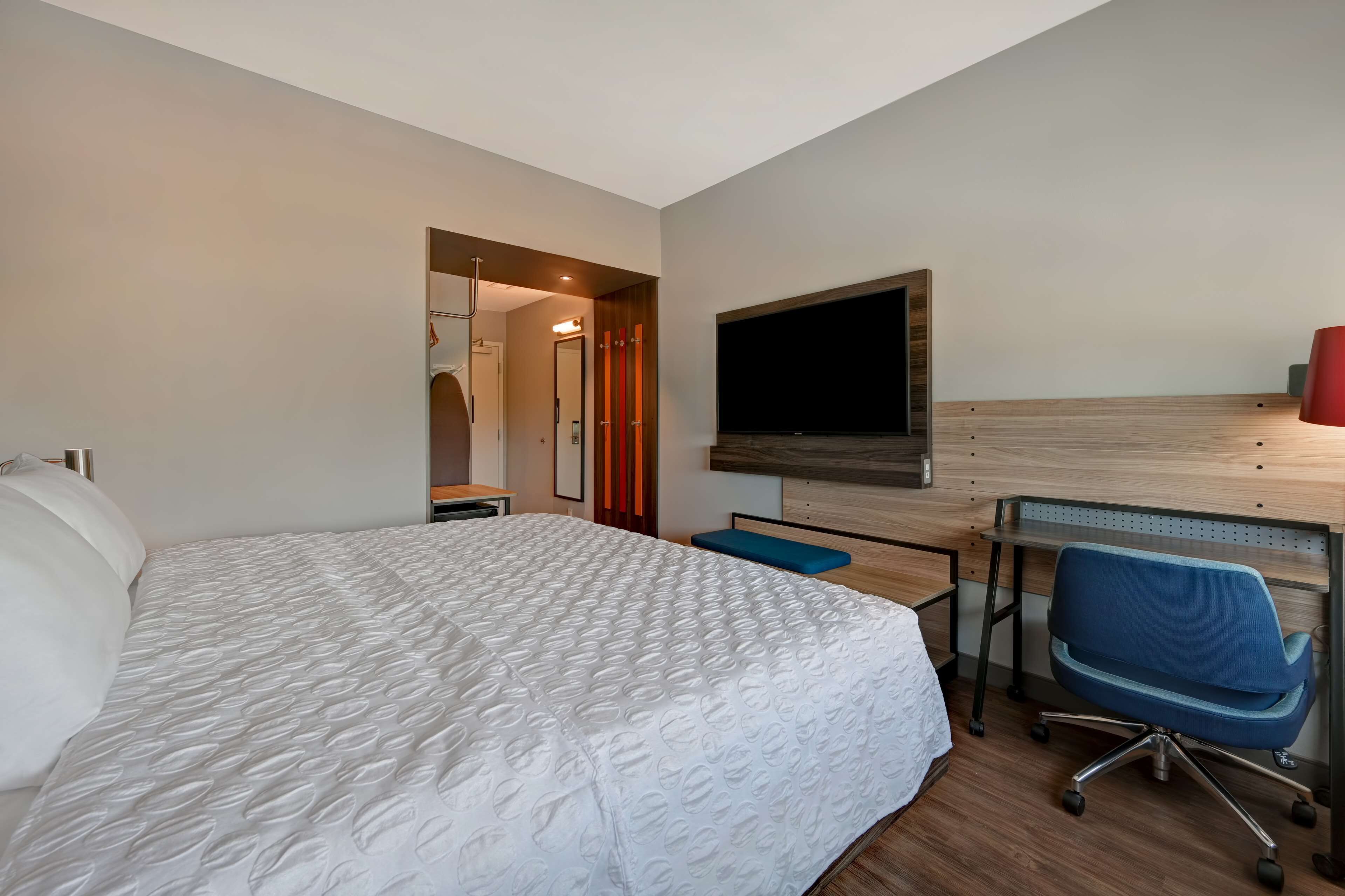Quarto de hotel moderno com uma cama grande, uma escrivaninha com cadeira, uma TV de tela plana na parede e piso de madeira.