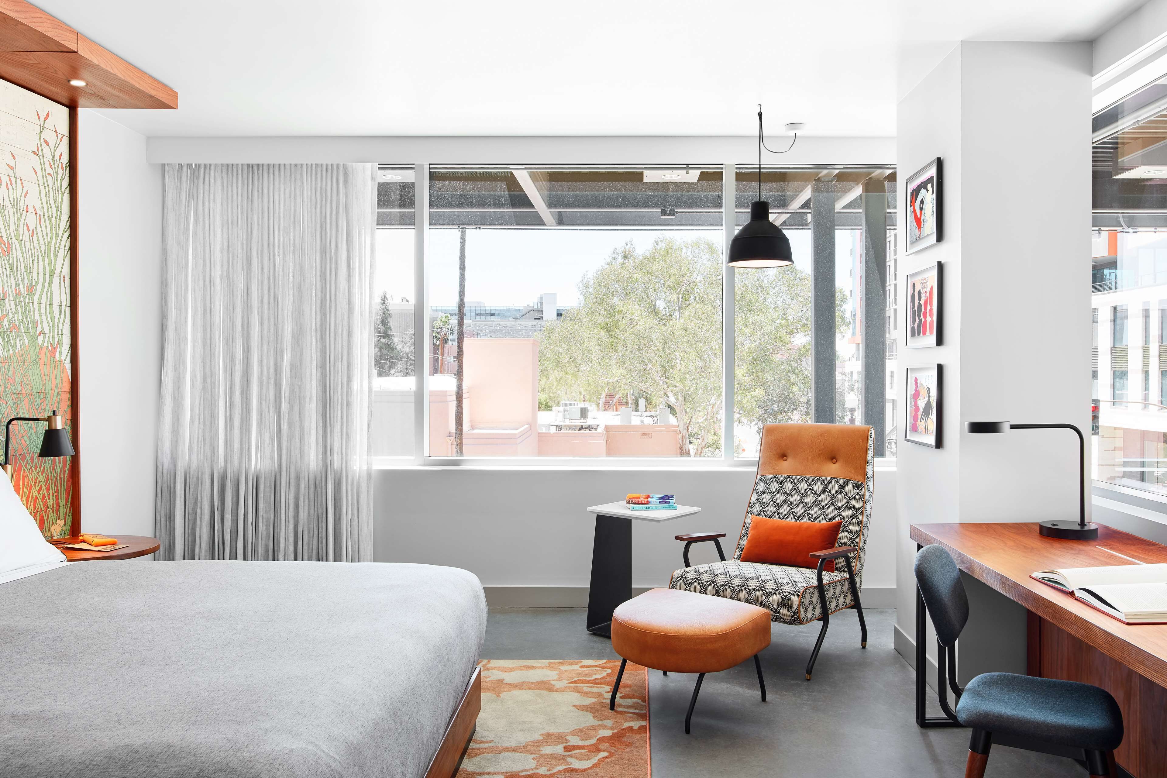 Quarto de hotel moderno com uma cama grande, uma escrivaninha, uma cadeira e vista da cidade através de amplas janelas.