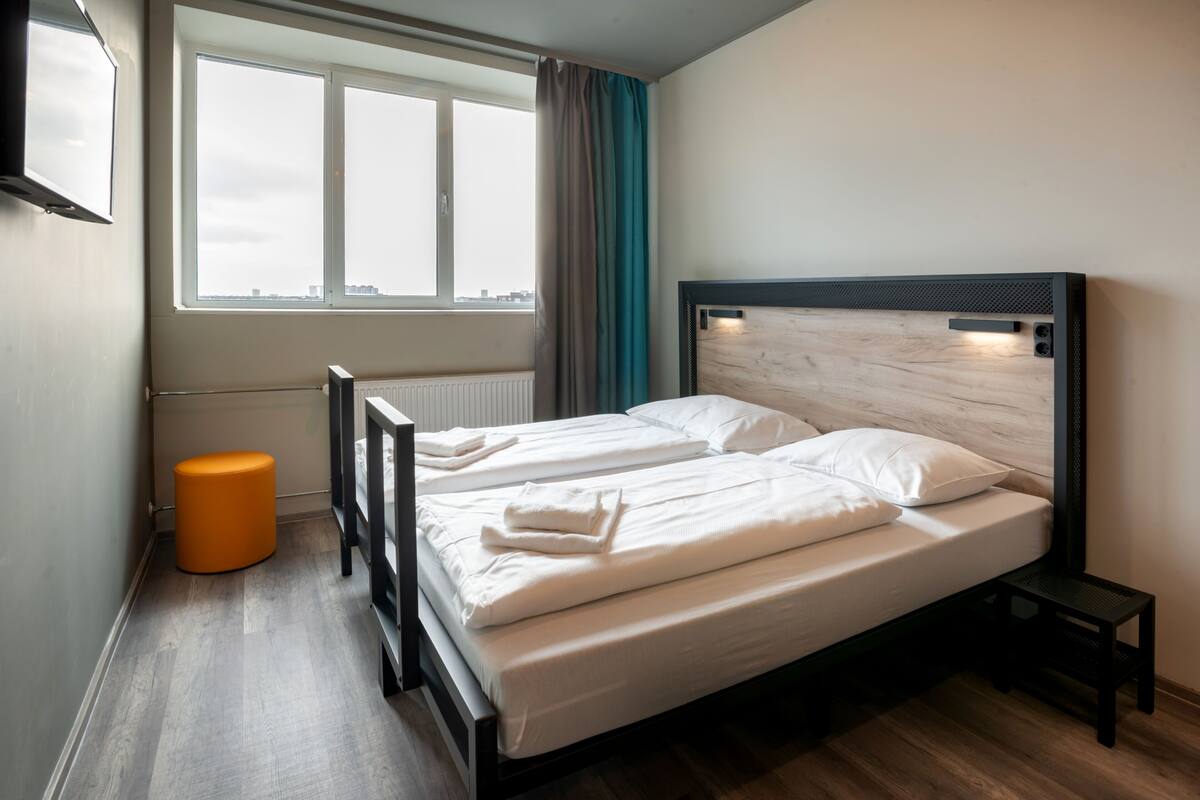 Quarto de hotel moderno com uma janela grande, piso de madeira, cama com roupa branca e um banquinho laranja pequeno.