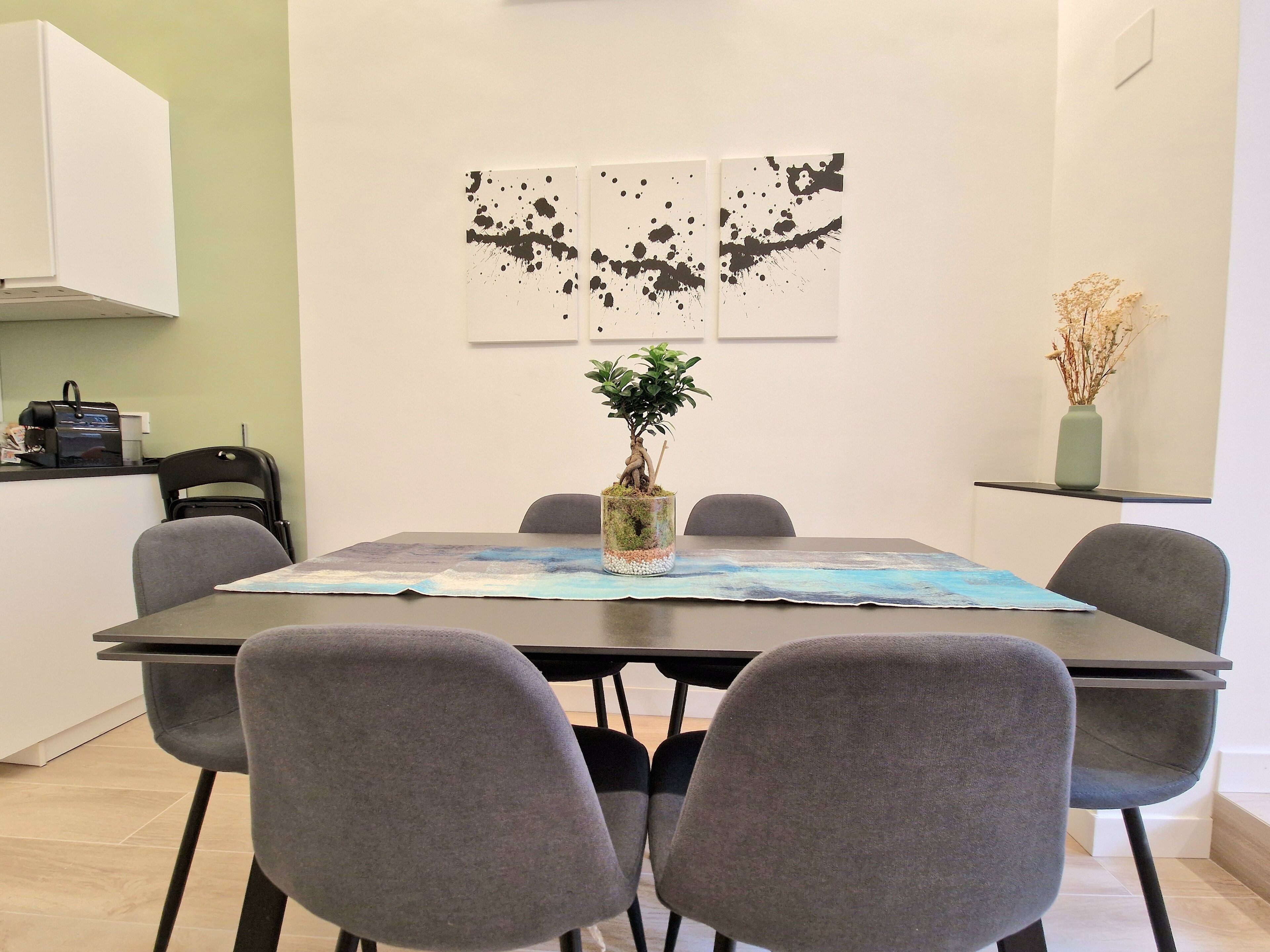 Uma sala de jantar moderna com uma mesa posta para seis pessoas, rodeada por cadeiras cinzentas. Há uma planta em vaso sobre a mesa, e obras de arte nas paredes.