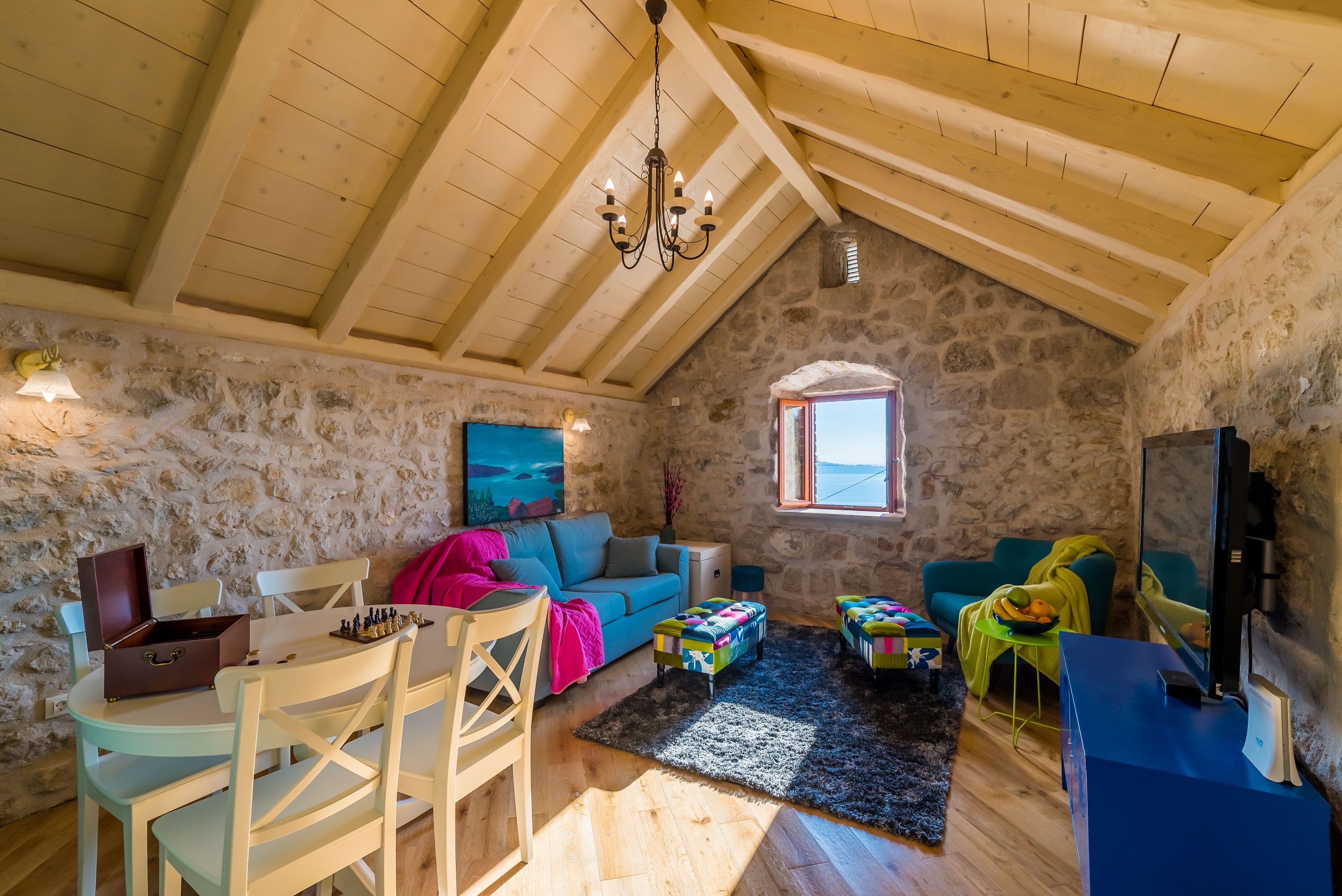 Um loft aconchegante com vigas de madeira aparentes, paredes de pedra, lareira de pedra, uma janela grande e um espaço de estar convidativo com um tabuleiro de xadrez.