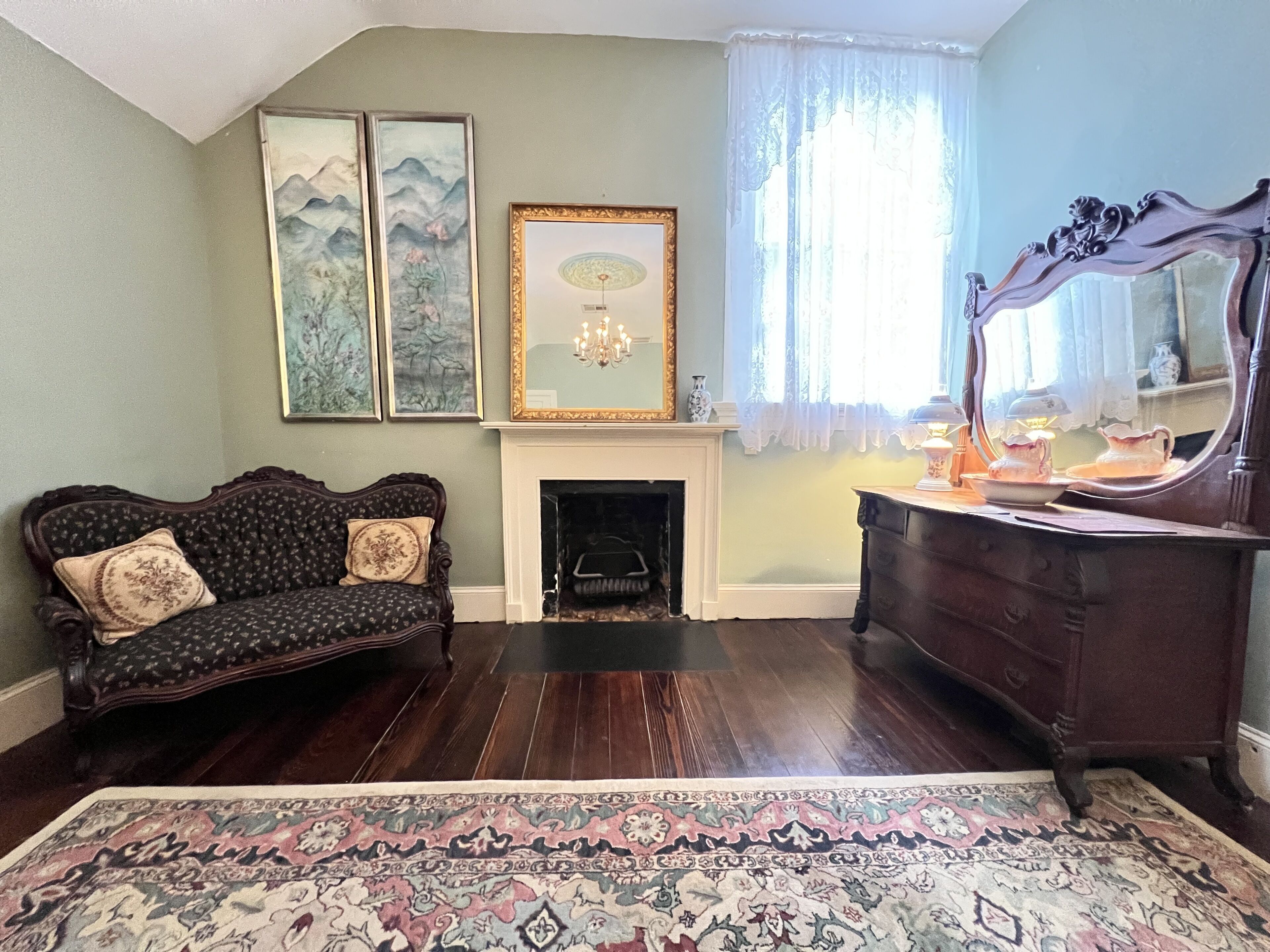 Um quarto com assoalho de madeira escura, um tapete estampado, um sofá escuro com almofadas decorativas, uma lareira, um espelho e duas obras de arte emolduradas na parede.