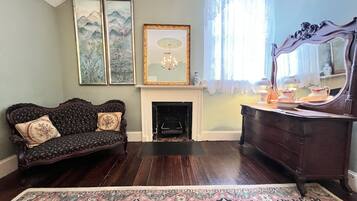 Um quarto com assoalho de madeira escura, um tapete estampado, um sofá escuro com almofadas decorativas, uma lareira, um espelho e duas obras de arte emolduradas na parede.