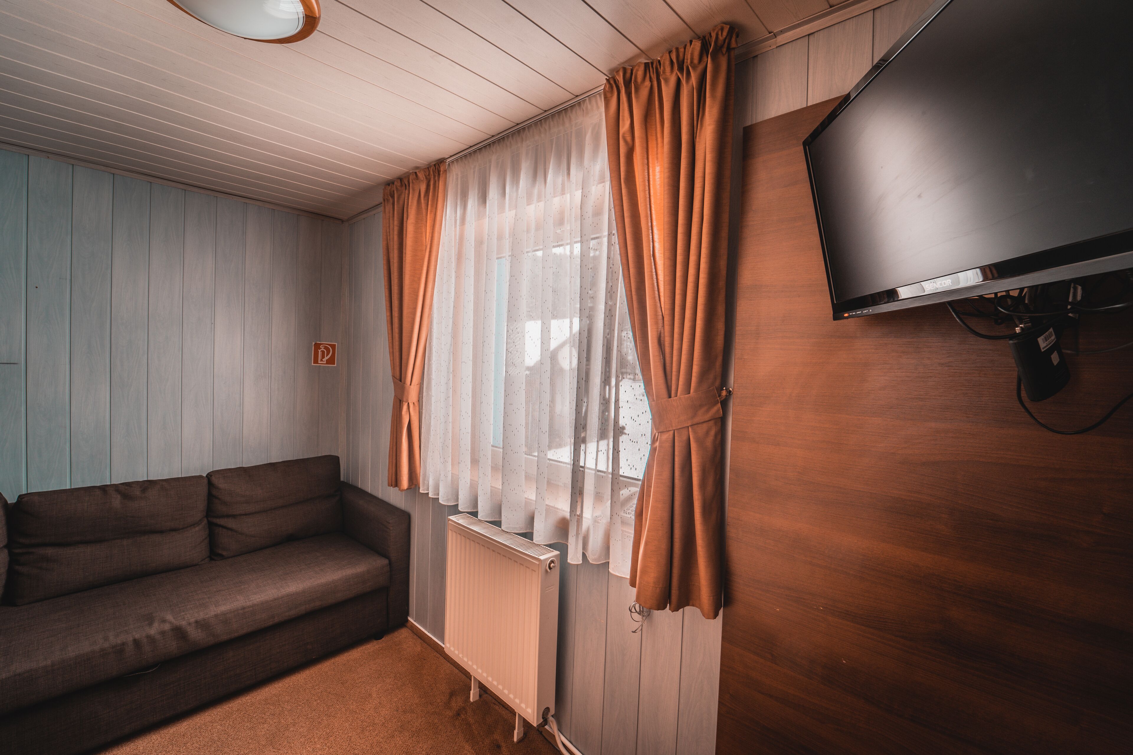 Quarto com sofá marrom, radiador, televisão fixada na parede e janela com cortinas leves.