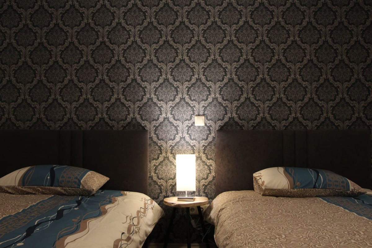 Quarto com duas camas, uma mesinha e uma luminária na parede.