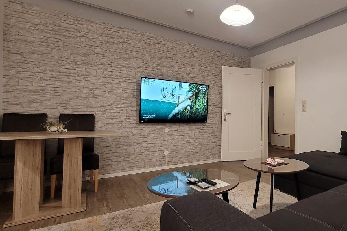 Uma sala de estar moderna com uma parede de pedra, uma TV de tela plana, um sofá escuro, uma mesa de centro de vidro e uma mesa de apoio de madeira.