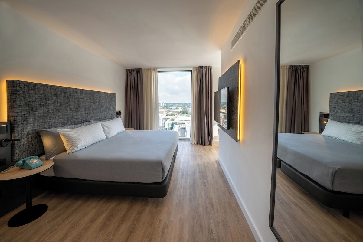 Quarto de hotel moderno com duas camas, uma janela grande com cortinas, televisão de tela plana e um espelho.