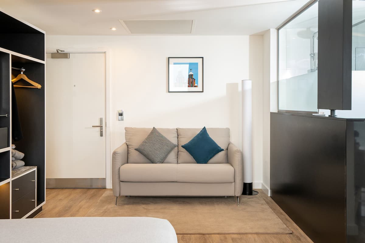 Quarto de hotel moderno com sofá bege, um quadro na parede e uma janela grande.