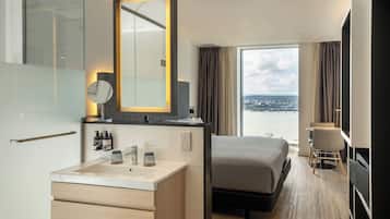 Quarto de hotel moderno com uma cama grande, banheiro com espelho e pia, e vista para a cidade.