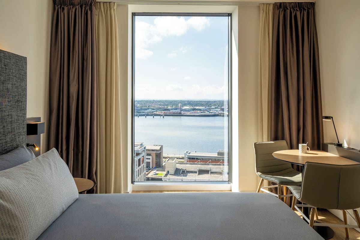 Quarto de hotel com uma cama grande, uma mesa redonda com cadeiras e vista para a cidade através de uma grande janela.