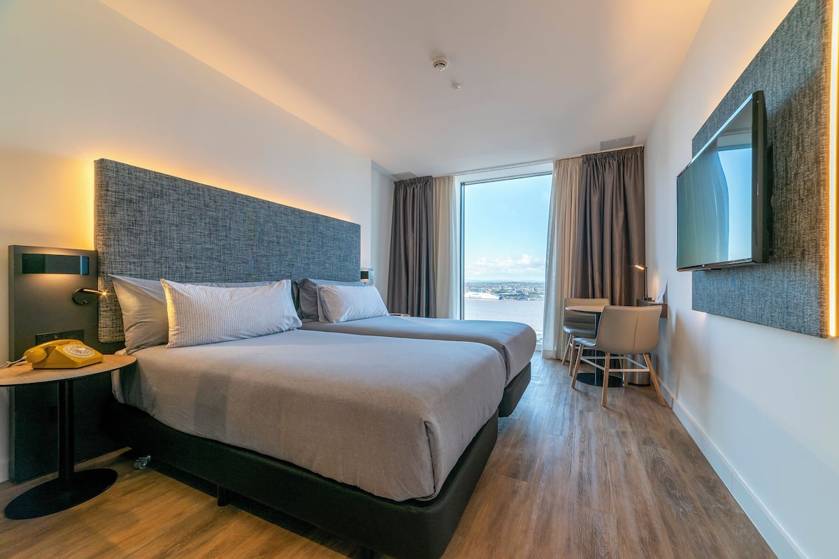Quarto de hotel com duas camas, uma escrivaninha com cadeira, televisão na parede e uma janela grande com cortinas.