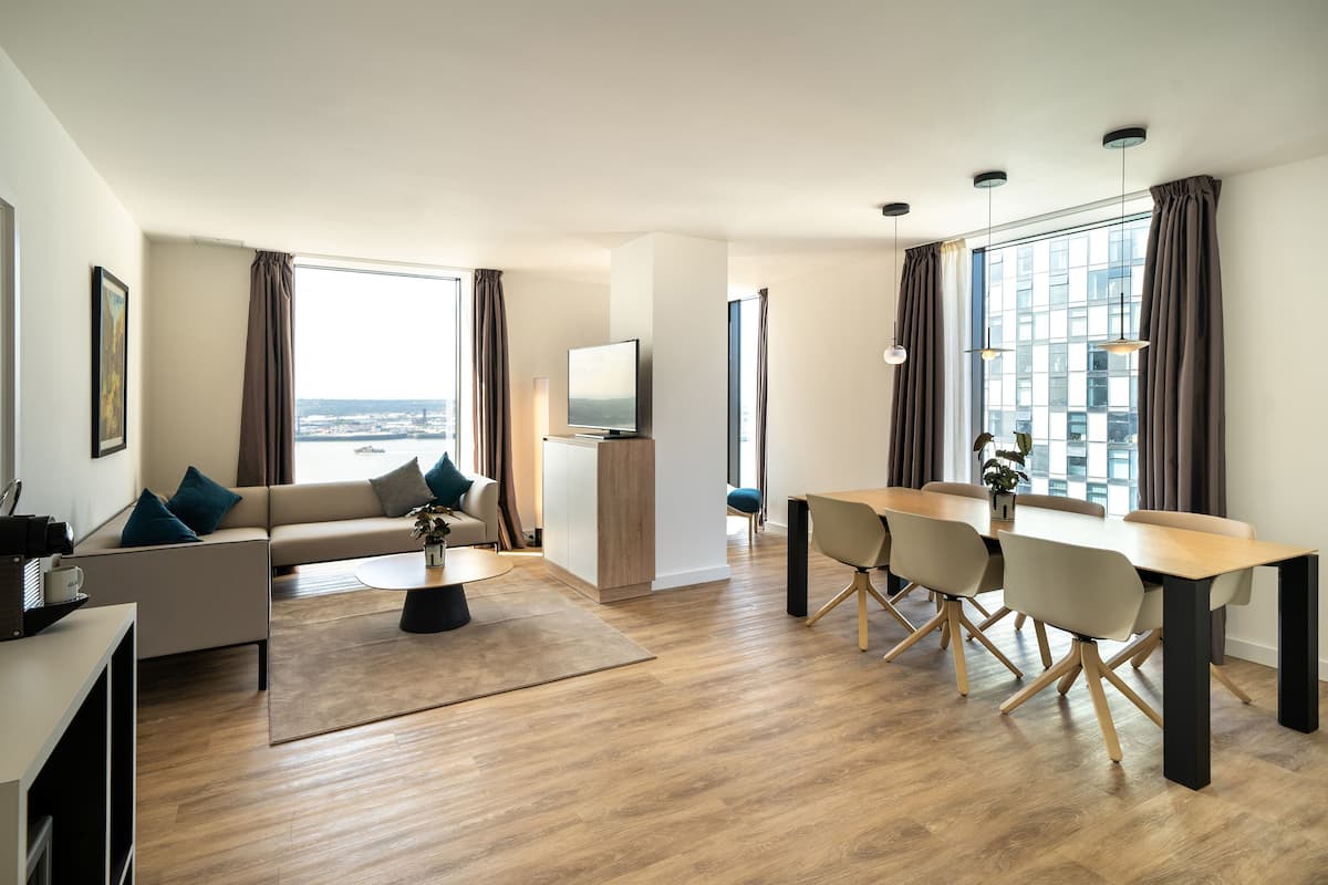 Uma sala de estar moderna com sofá, mesa de centro e mesa de jantar. Há uma televisão sobre um suporte e grandes janelas com cortinas.