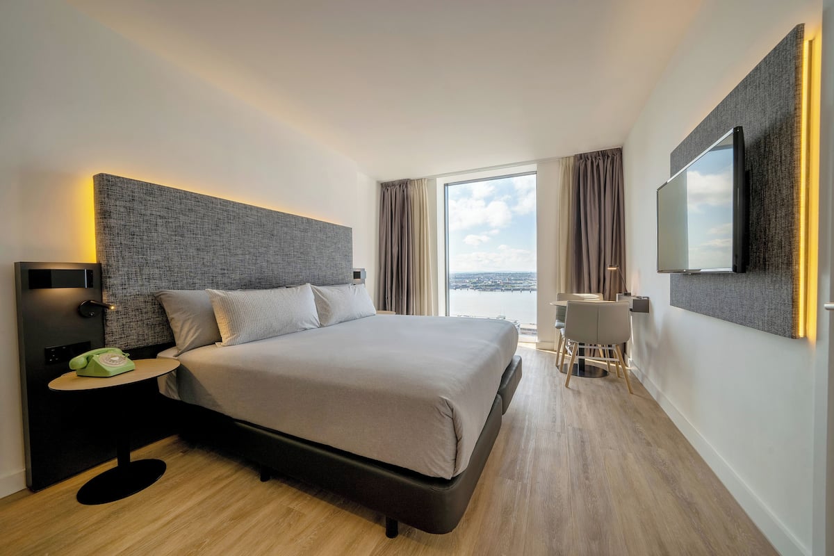 Quarto de hotel moderno com uma cama grande, uma mesa de trabalho, uma cadeira, televisão de tela plana na parede e vista para a cidade pela janela.