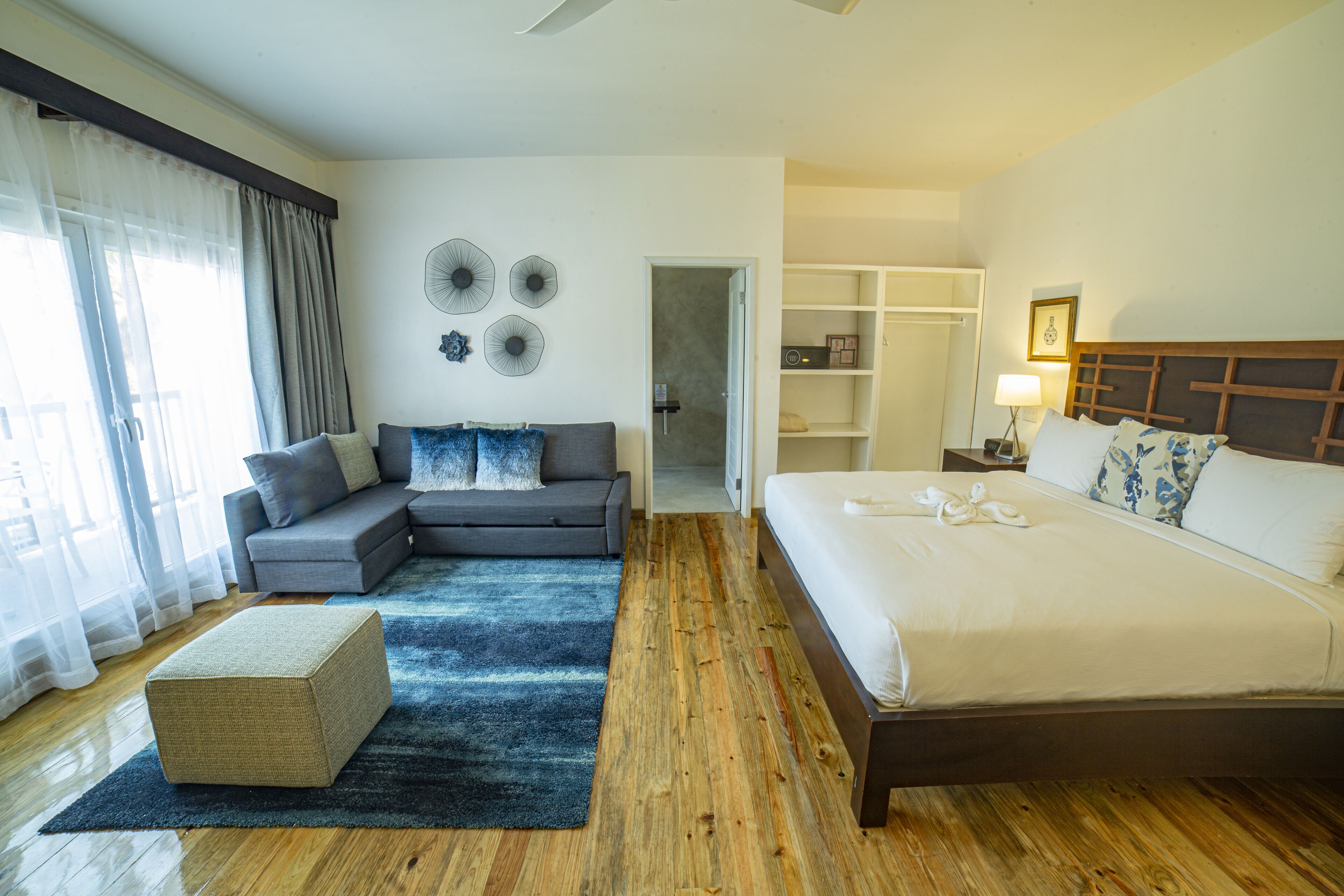 Quarto de hotel moderno com uma cama grande, um sofá, um pequeno banquinho e chão de madeira, além de um closet espaçoso.