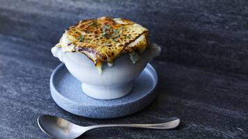 Uma tigela de sopa com uma cobertura dourada, sobre uma superfície escura e texturizada, com uma colher ao lado.