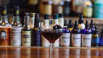 Um bar com um copo de bebida escura, decorado com uma baga, e várias garrafas de bebidas alcoólicas ao redor.