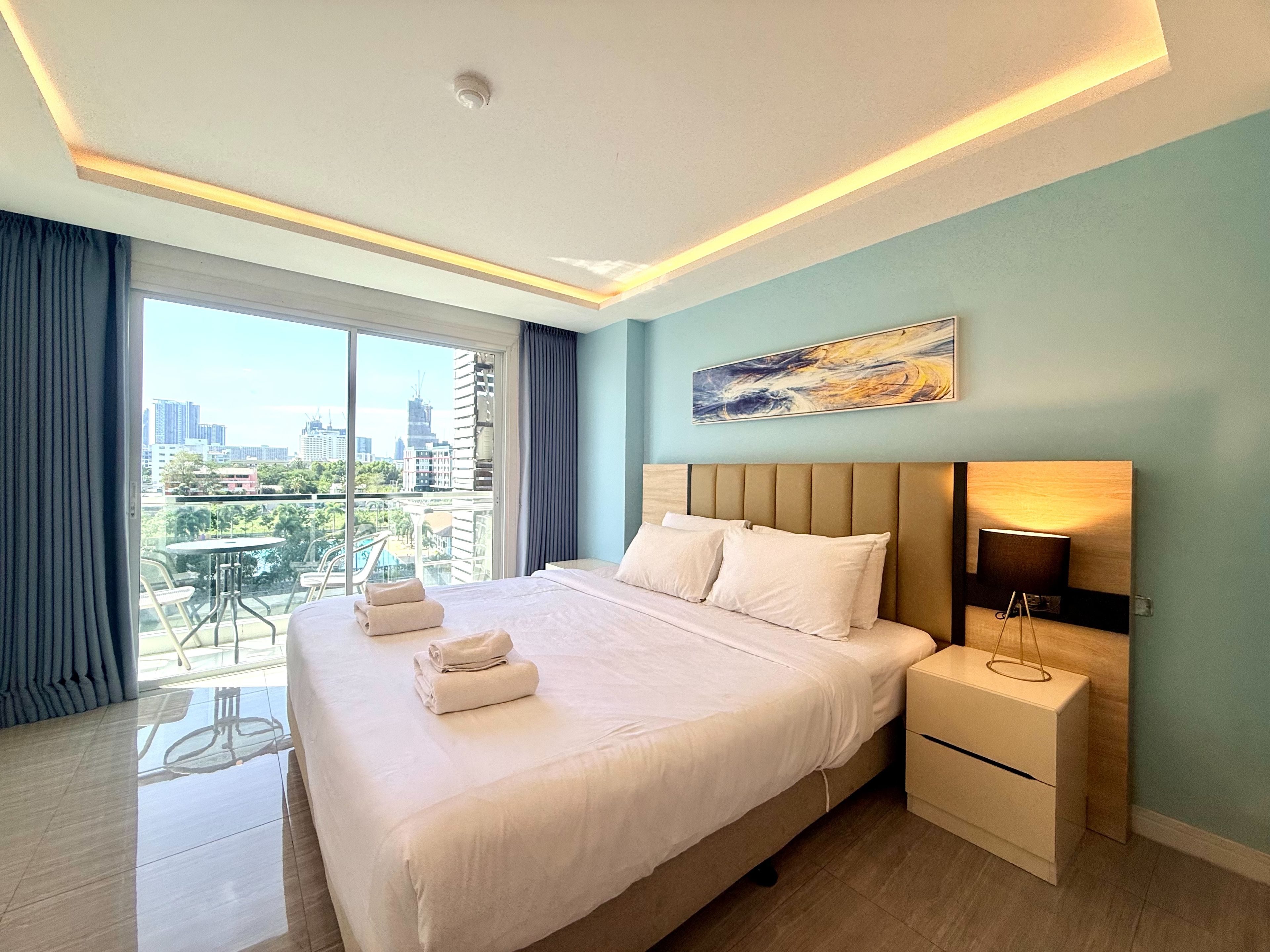 Quarto de hotel com uma cama grande, mesa de cabeceira, vista para a cidade e varanda com móveis de exterior.