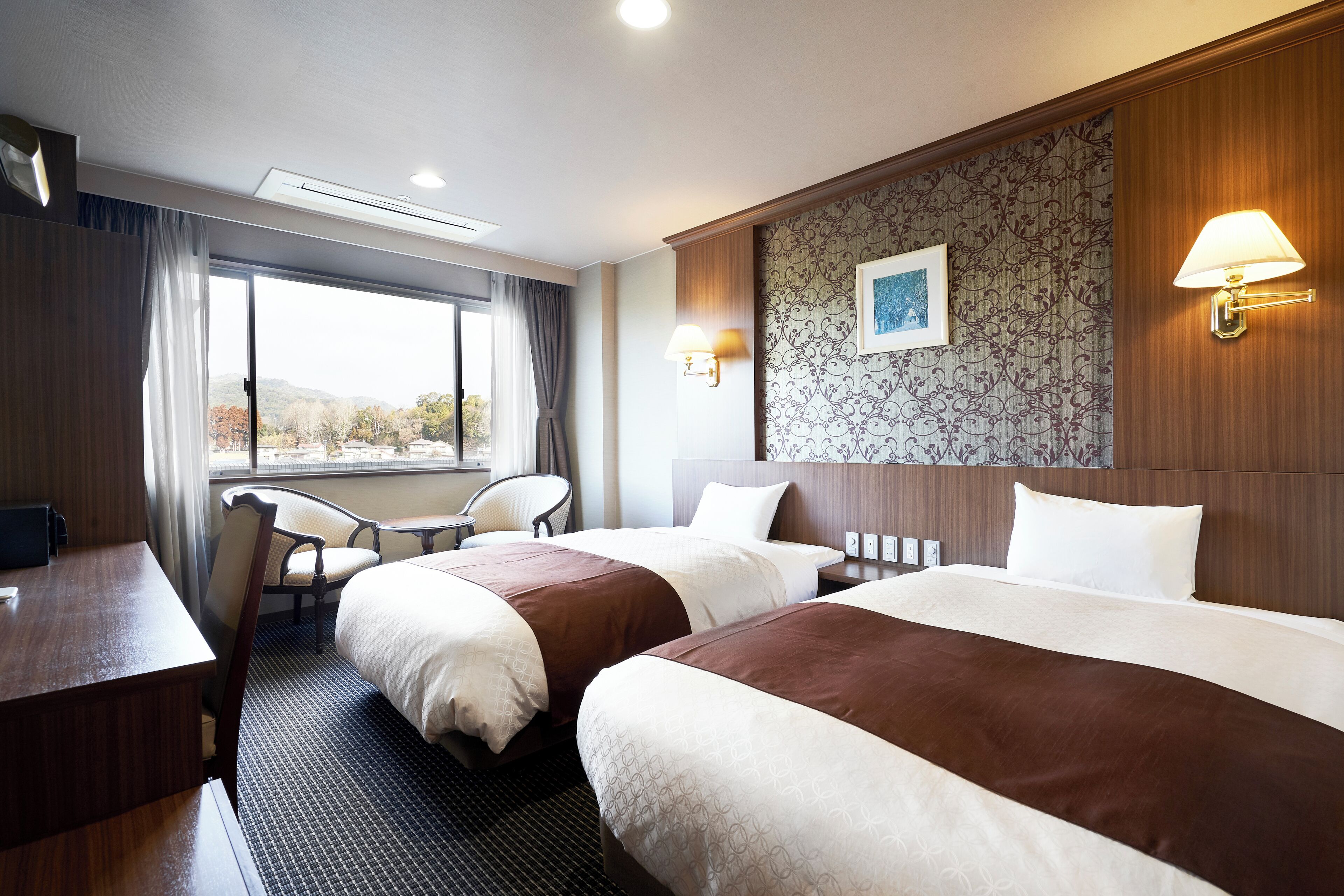 Quarto de hotel com duas camas, uma escrivaninha, uma cadeira e janela com vista para árvores.