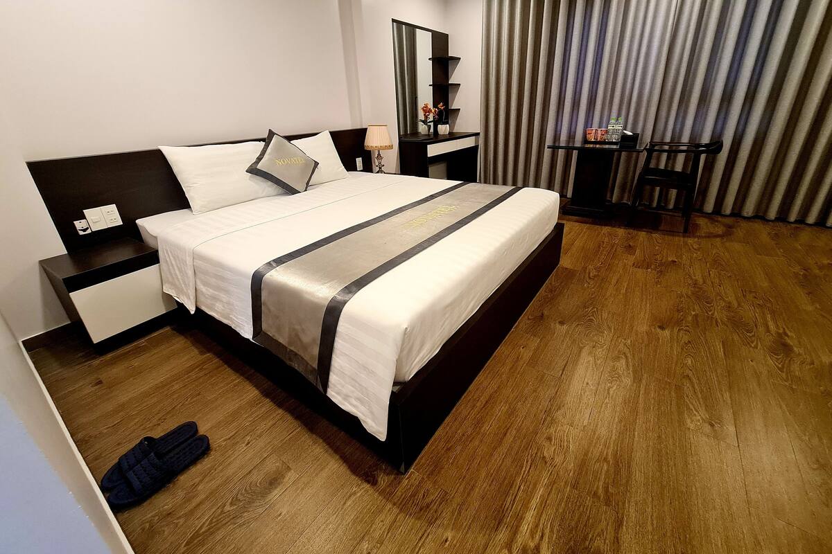 Quarto de hotel com uma cama grande, uma mesa de cabeceira, um abajur, uma cadeira e uma cortina.