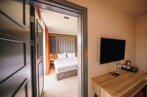 Quarto de hotel com cama, mesa de cabeceira, televisão e escrivaninha de madeira.