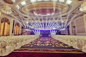 Um salão imponente com um tapete longo e ricamente decorado, fileiras de cadeiras brancas e um palco com fundo azul.