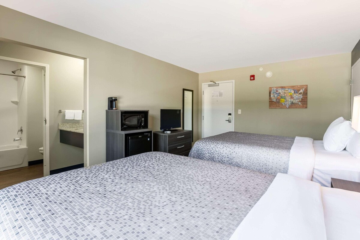 Quarto de hotel com duas camas, uma mesa de trabalho com computador, micro-ondas e chuveiro.