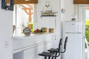 Uma cozinha com bancada branca, decoração de um veleiro pendurado, uma pequena prateleira com cestos e uma tigela decorativa, além de uma geladeira.