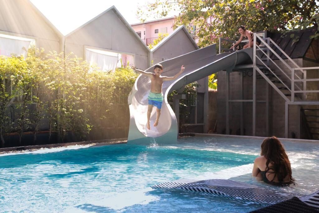 Uma piscina com escorrega, uma pessoa escorregando e outra sentada à beira da piscina.