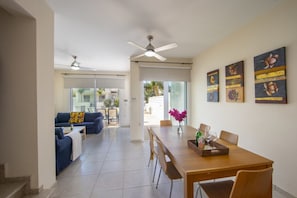 Uma sala de estar moderna com uma mesa de jantar, cadeiras e um sofá. Há ventiladores no teto, uma janela grande e obras de arte na parede.