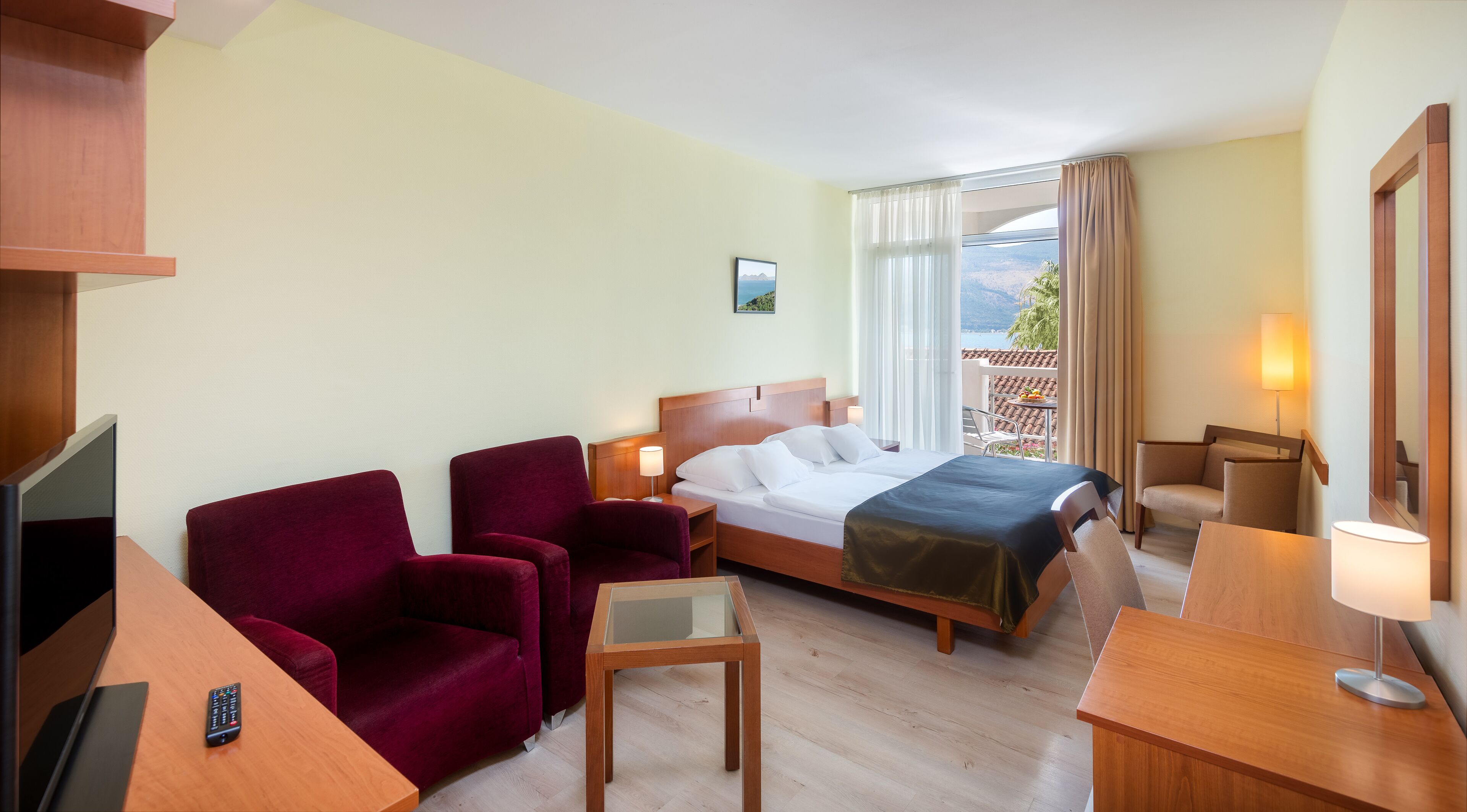 Quarto de hotel com cama, duas poltronas vermelhas, uma mesa pequena, televisão e varanda com vista.