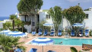Uma área junto à piscina com espreguiçadeiras, guarda-sóis e uma grande árvore.