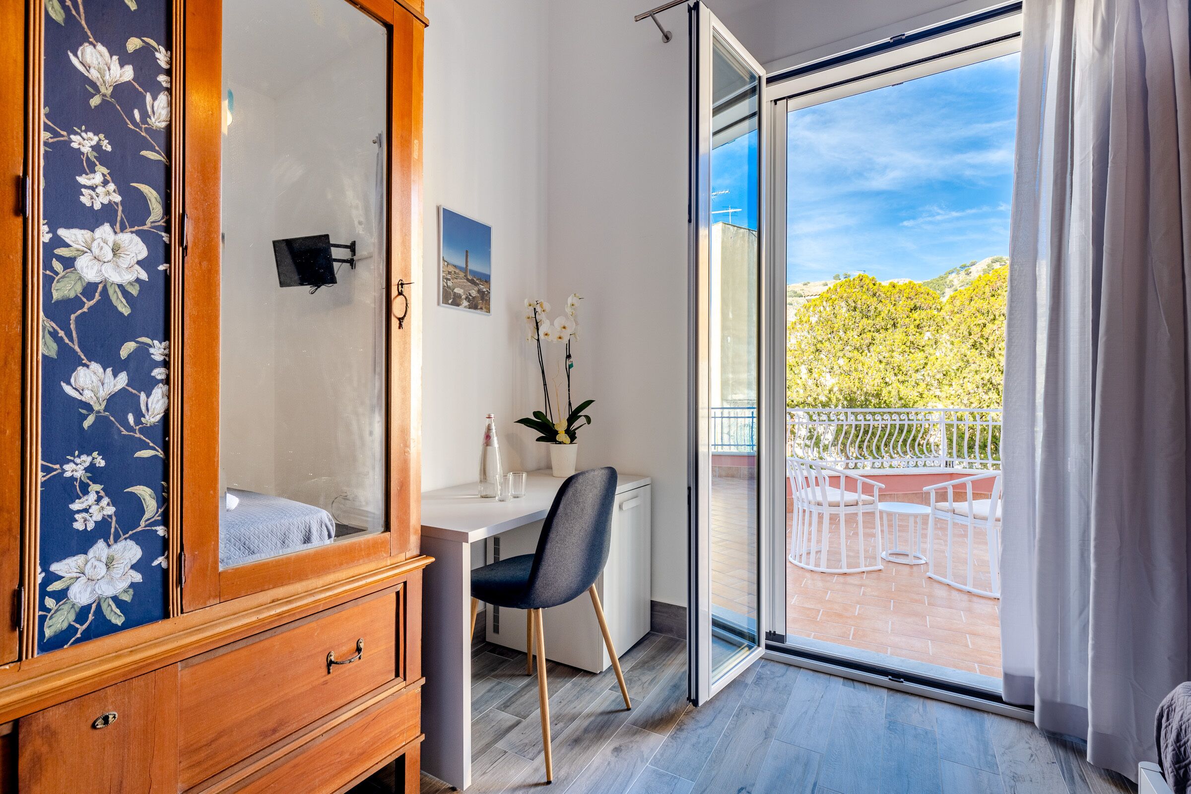 Quarto com armário de madeira, uma escrivaninha, cadeira, planta em vaso e vista para uma varanda com mobiliário exterior.