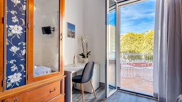 Quarto com armário de madeira, uma escrivaninha, cadeira, planta em vaso e vista para uma varanda com mobiliário exterior.