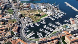 Vista aérea de uma cidade costeira com um porto de iates, edifícios e uma montanha imponente ao fundo.