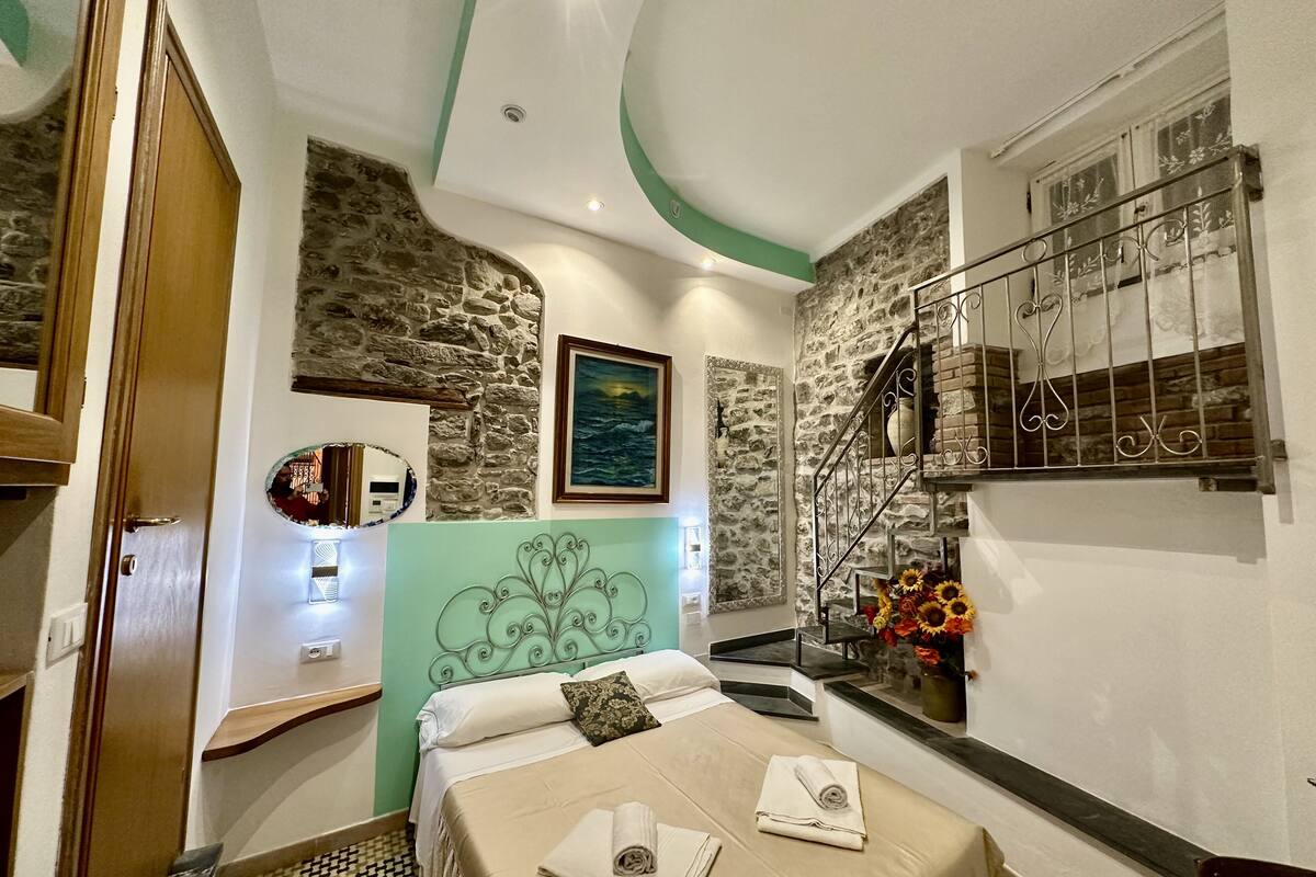 Quarto com parede de pedra, cama com toalhas dobradas, espelho e escada com corrimão de metal.