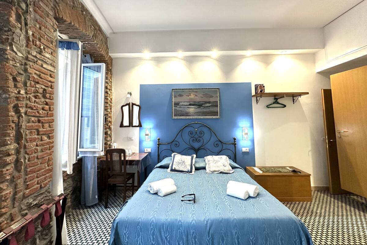 Quarto com parede de tijolos, cama com colcha azul, escrivaninha com cadeira e guarda-roupa de madeira.