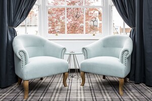 Duas poltronas revestidas em azul claro, com pernas de madeira e detalhes decorativos em tachas, dispostas sobre um piso xadrez, com uma pequena mesa entre elas.