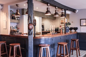 Uma área de bar com banquetas de madeira, luminárias pendentes em cobre e diversas garrafas e copos em prateleiras.