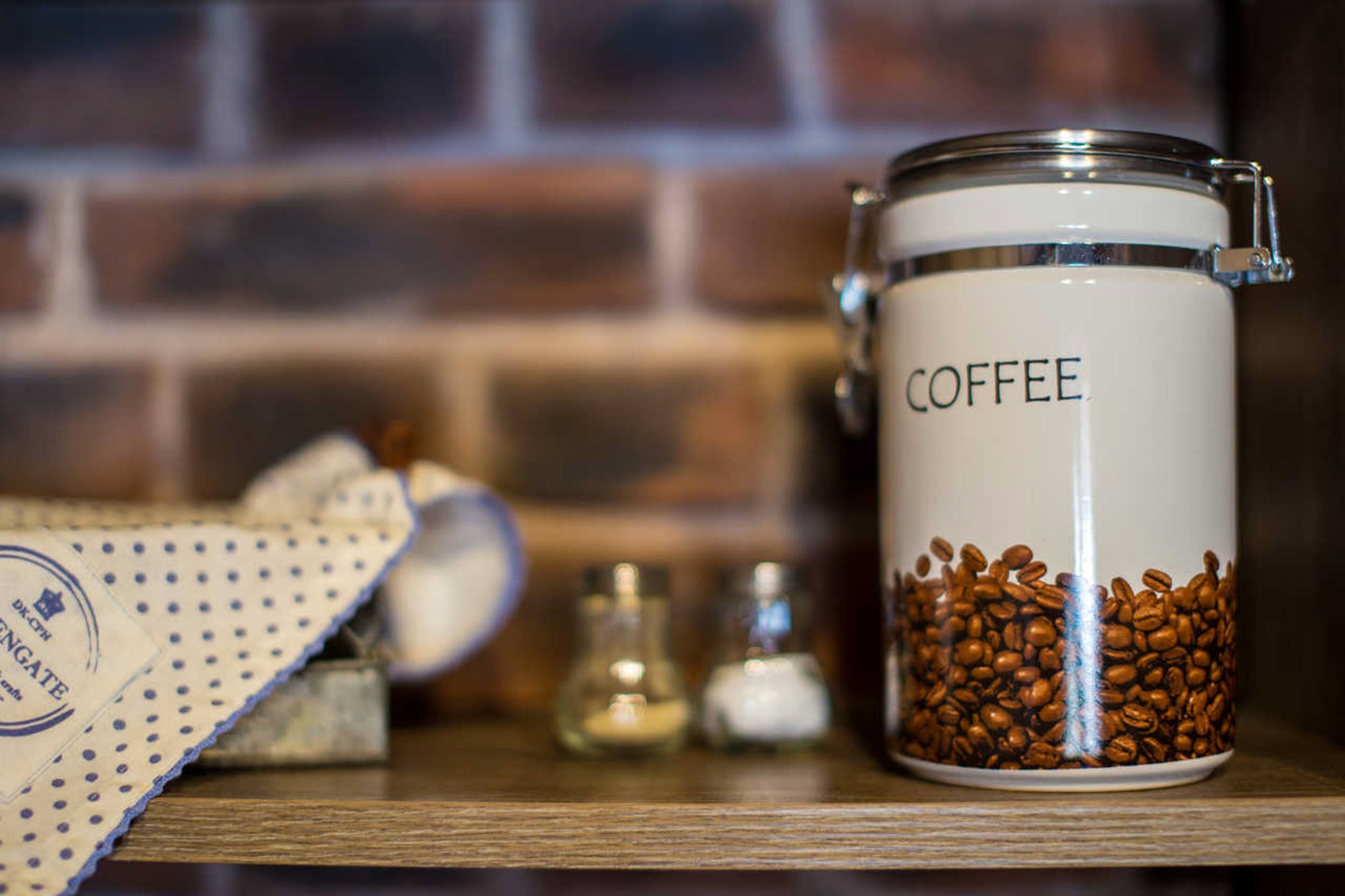 Recipiente de café com a inscrição "COFFEE" cheio de grãos de café, uma guardanapo de pano e saleiro e pimenteiro sobre uma superfície de madeira.