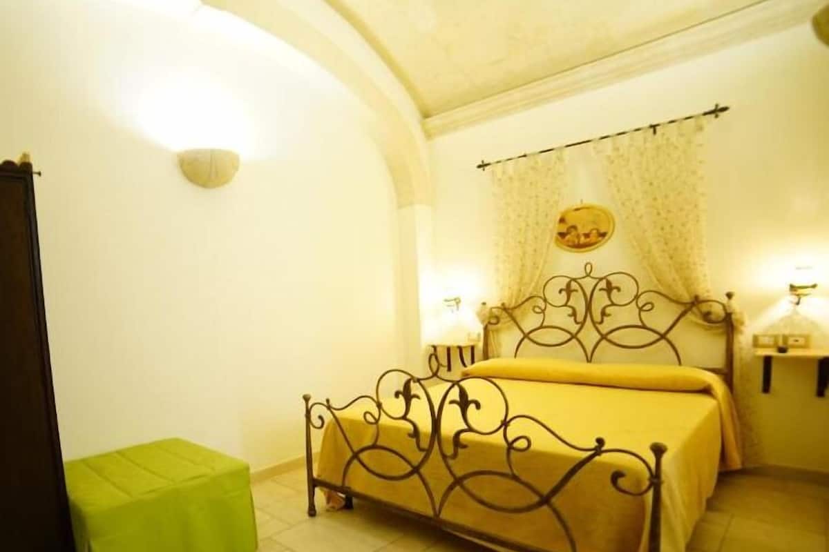 Quarto com uma cama, um banquinho verde, duas luminárias de parede e uma peça decorativa na parede.
