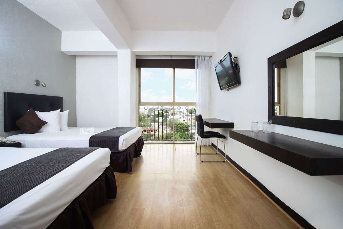 Quarto de hotel com duas camas, uma escrivaninha, uma cadeira, televisão e varanda com vista.