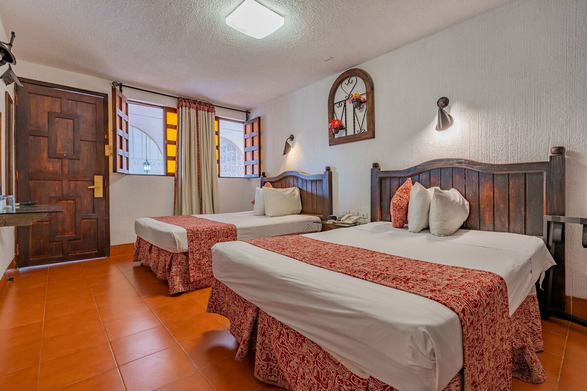 Quarto de hotel com duas camas, uma porta de madeira, janela com cortinas, espelho fixado na parede e uma pequena mesa com telefone.
