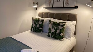Cama bem arrumada com lençóis brancos, duas almofadas com padrão de folhas, uma luminária de cabeceira e uma prateleira na parede com arte emoldurada e uma placa com a inscrição 'STAY'.
